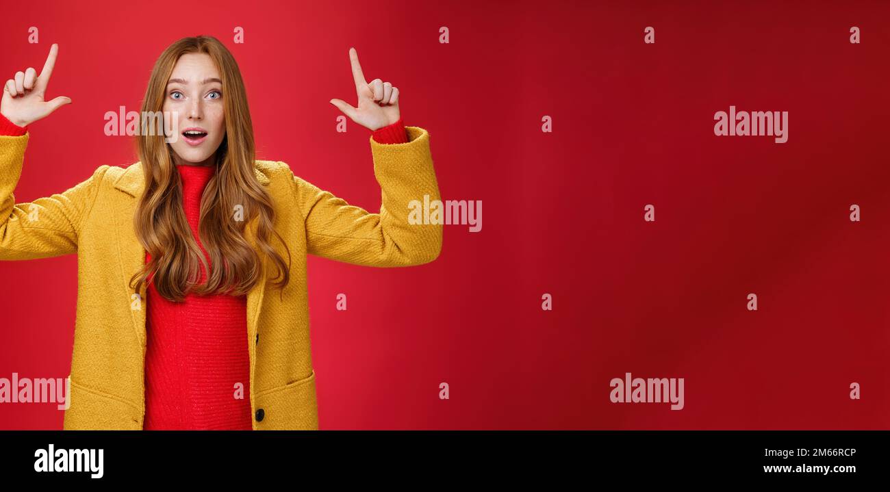 Surprised redhead girl in yellow coat open mouth impressed and thrilled as raising hands and ...