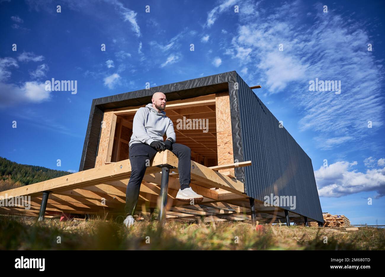 Male carpenter building wooden frame house on pile foundation. Bald man sitting on terrace on ...