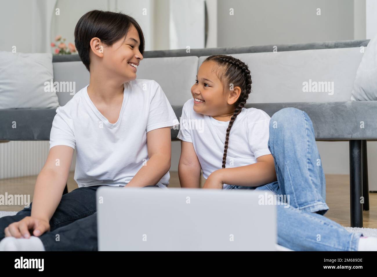 Positive asian siblings looking at each other near blurred laptop at ...