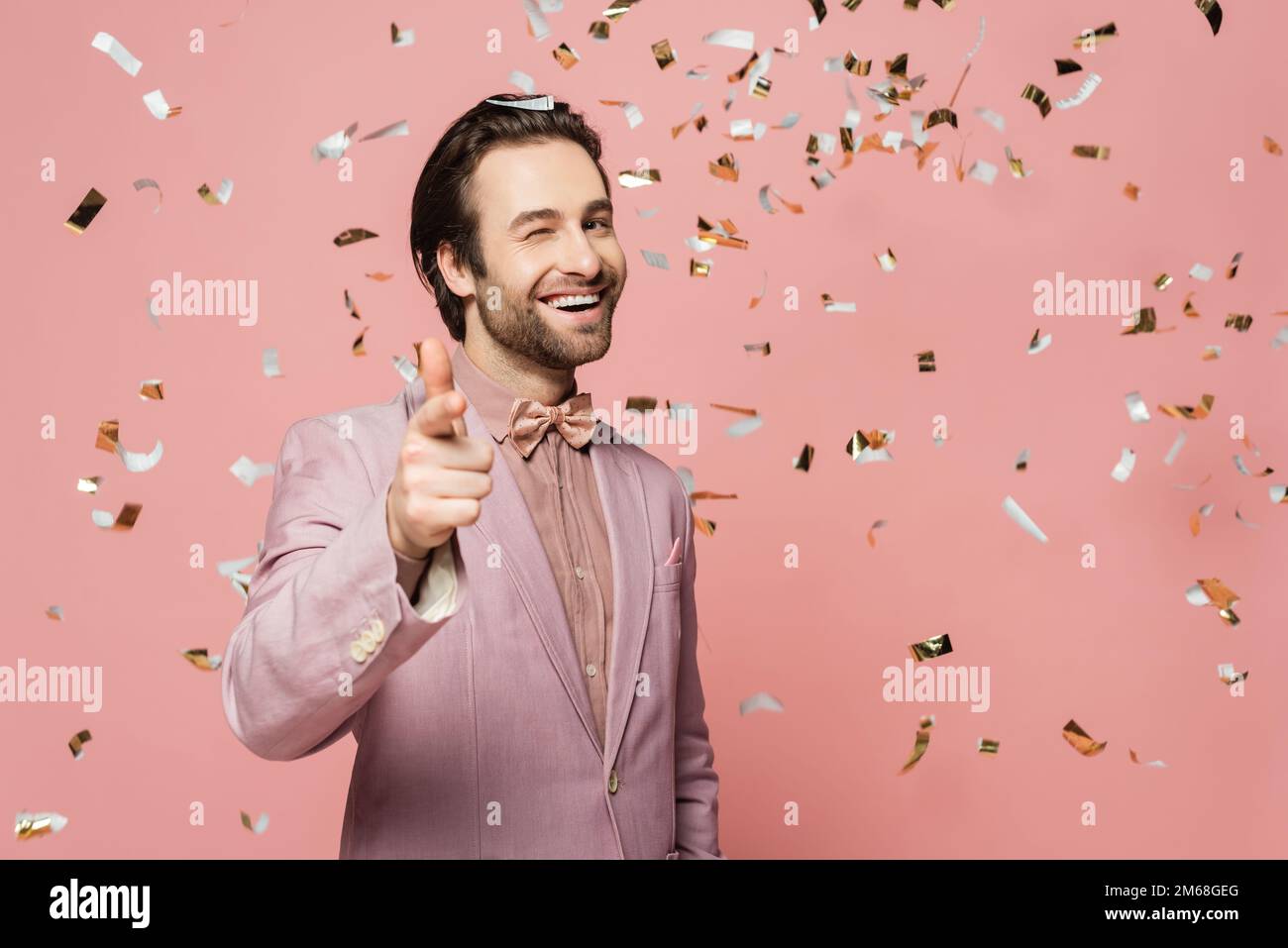 Smiling host of event pointing at camera under falling confetti on pink ...