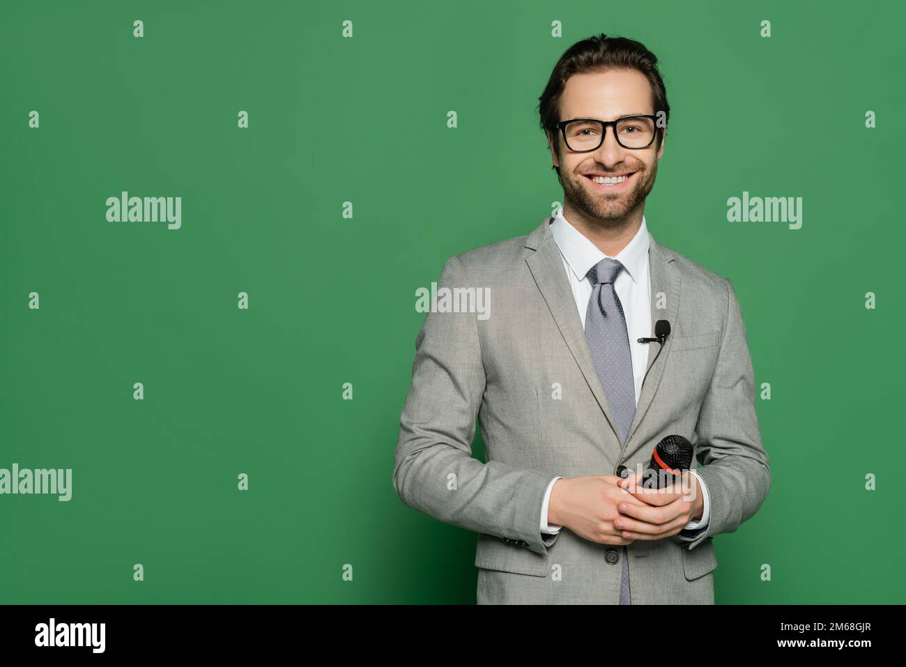 cheerful news anchor in suit and eyeglasses holding microphone on green ...