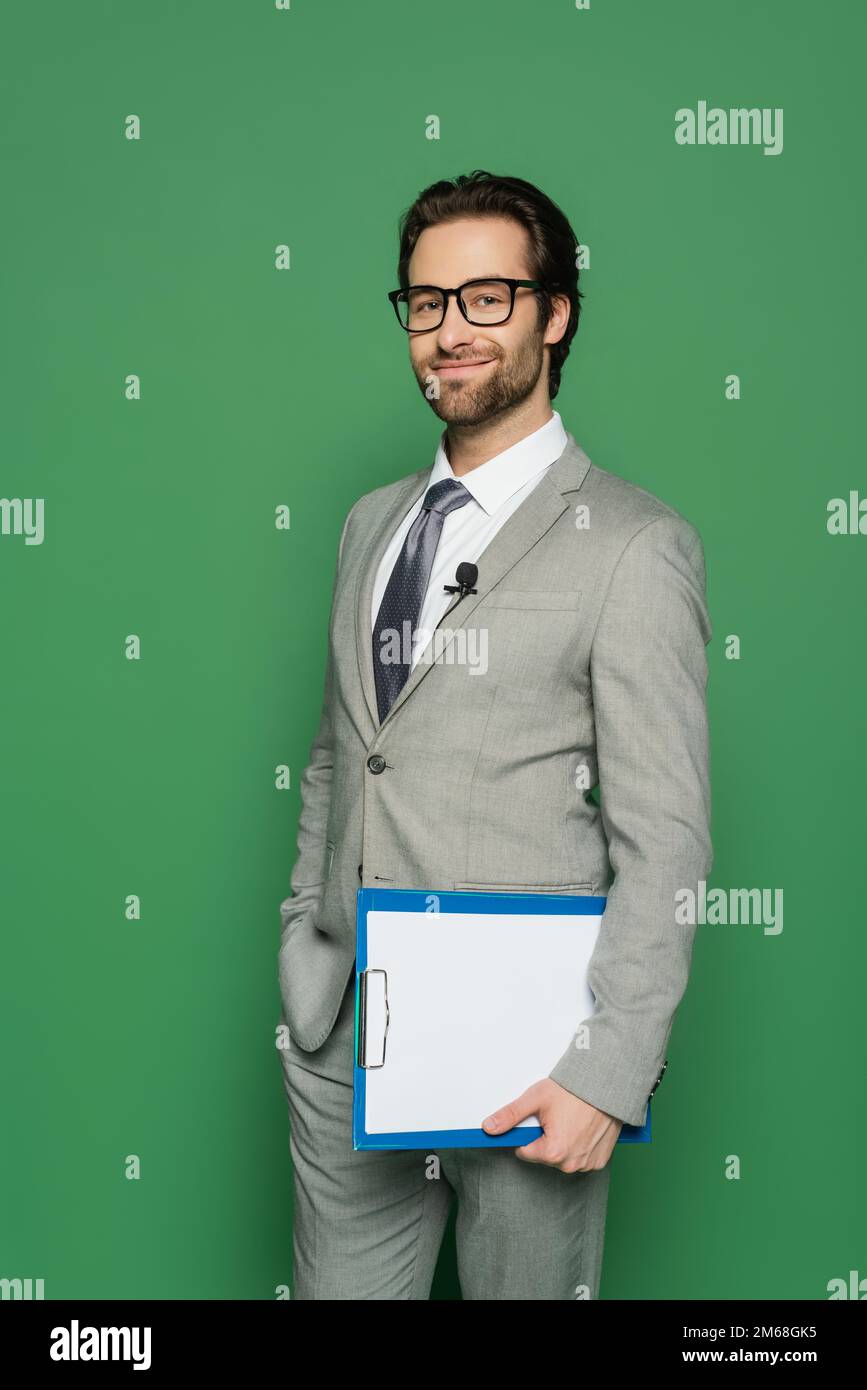 happy news anchor in suit and eyeglasses holding clipboard while ...