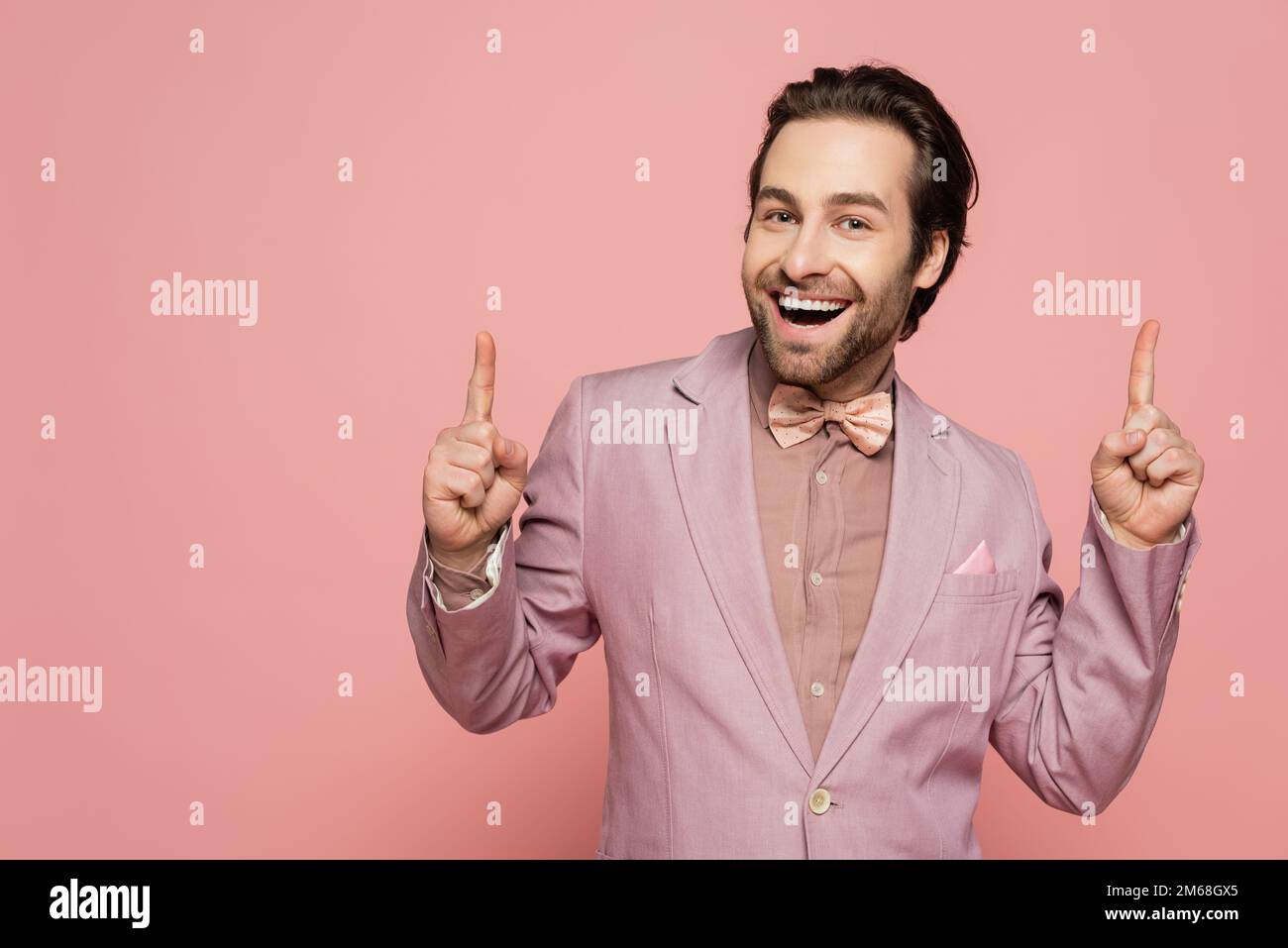 cheerful and young showman in suit with bow tie pointing up with ...