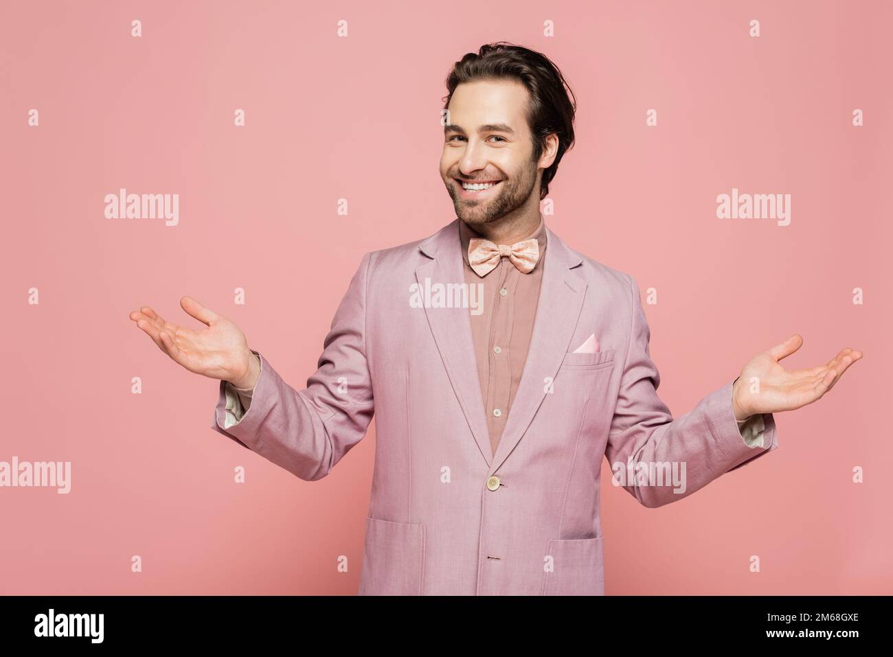 happy and young showman in suit with bow tie gesturing isolated on pink ...