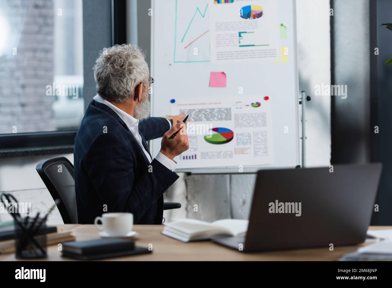 Grey haired businessman looking at blurred flip chart near laptop and coffee in office,stock ...