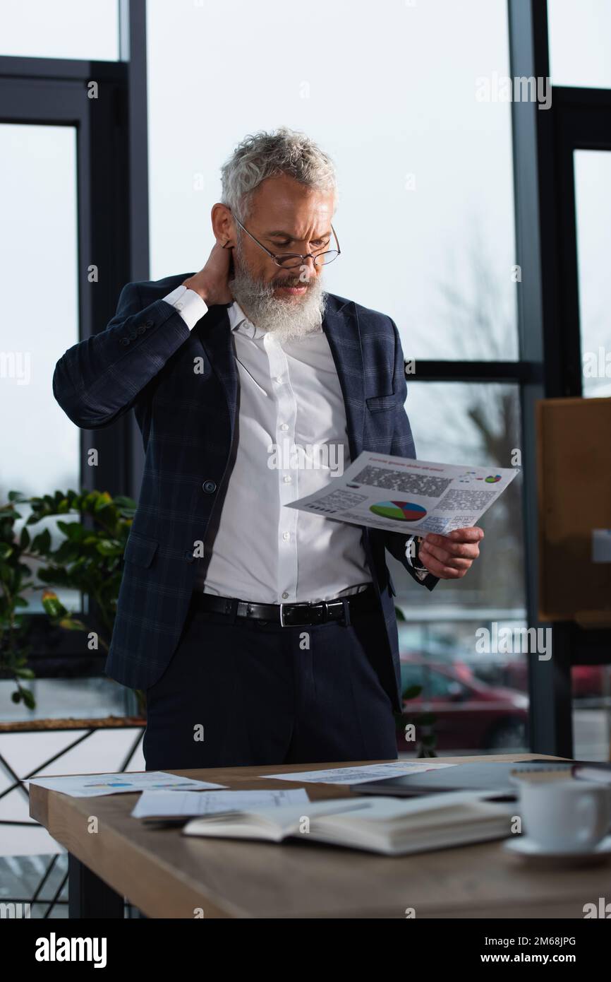 Middle aged manager holding paper with charts near working table in ...