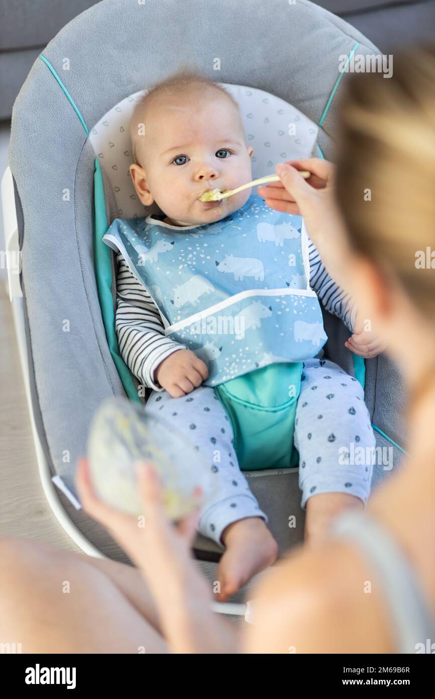 Mother spoon feeding her baby boy child in baby chair with fruit puree ...