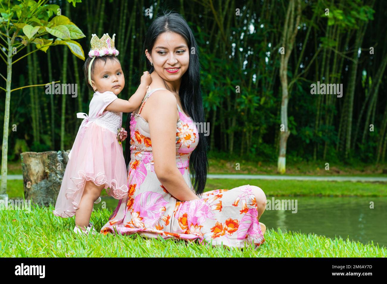 latina mother with her beautiful brunette baby, sitting by a lake next ...