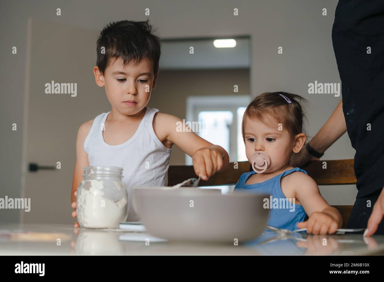 Two kids with their mother preparing dough for cookies together, adding ...