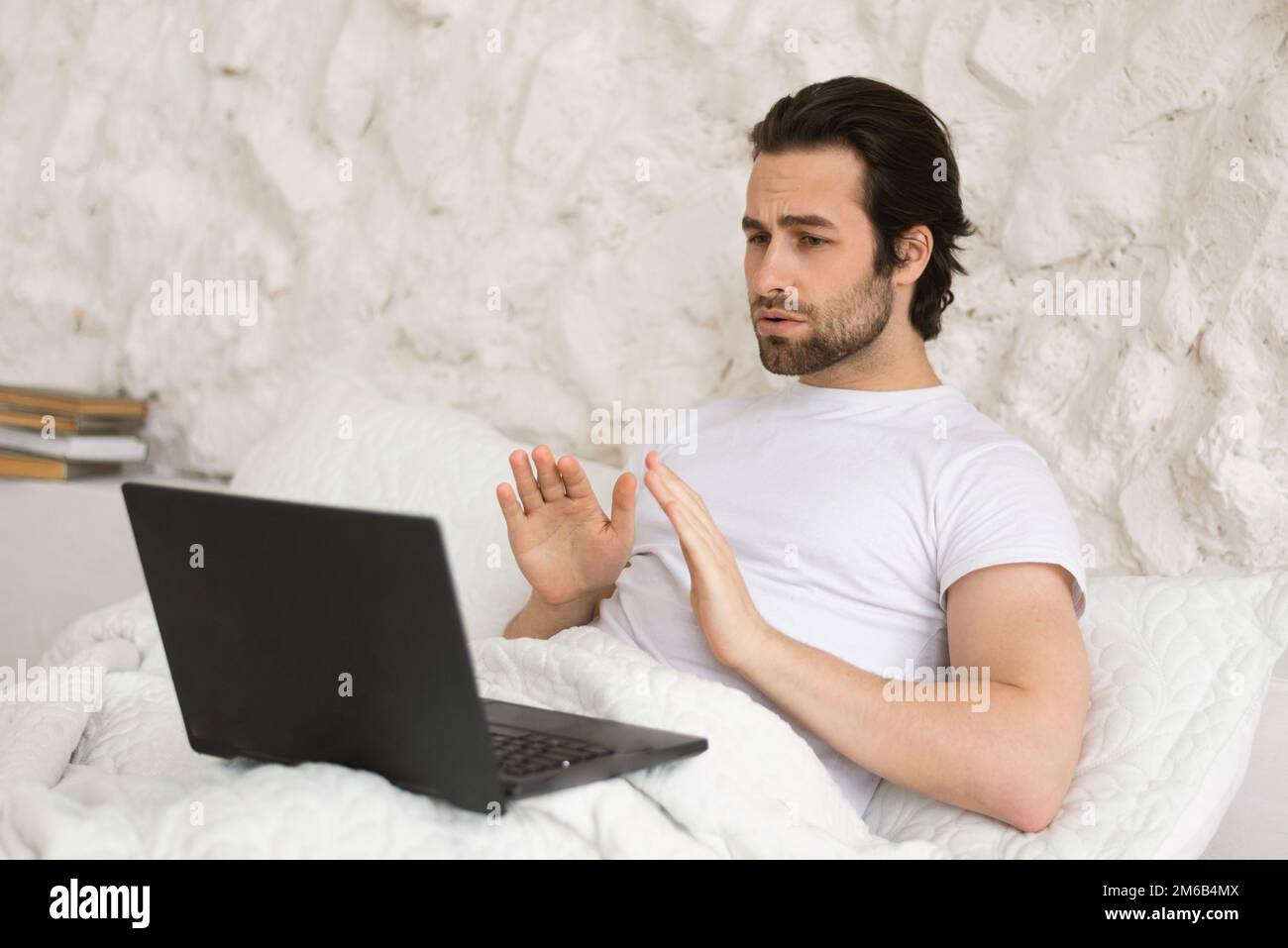 Sad serious young caucasian guy sit on bed, looks at computer, has ...