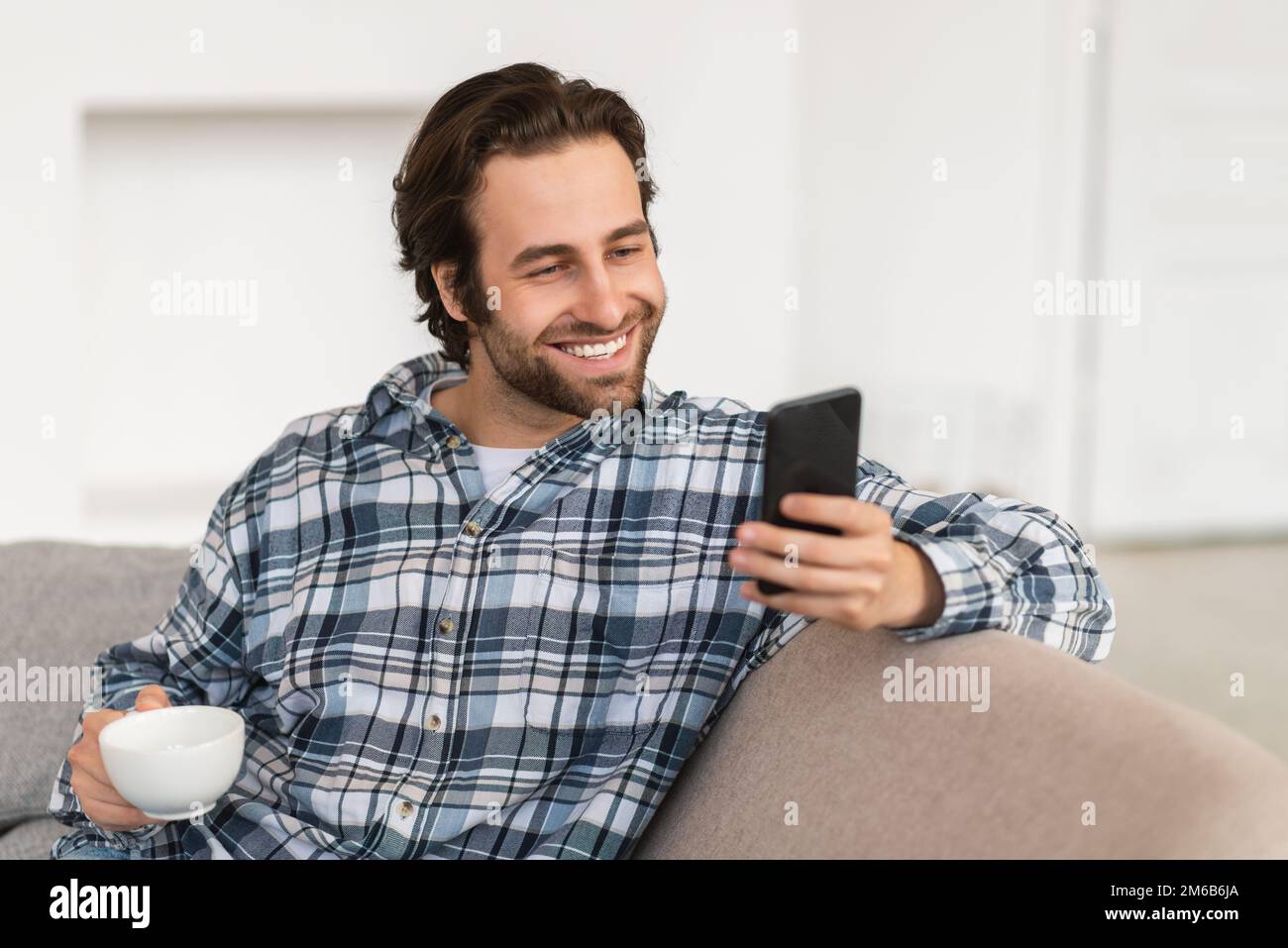 Glad young caucasian male sits on sofa, enjoys cup of coffee and ...