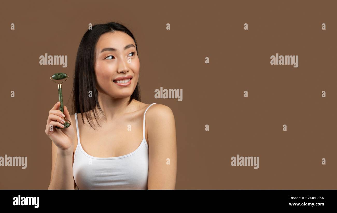 Pretty korean lady using greenstone jade facial roller, looking aside ...