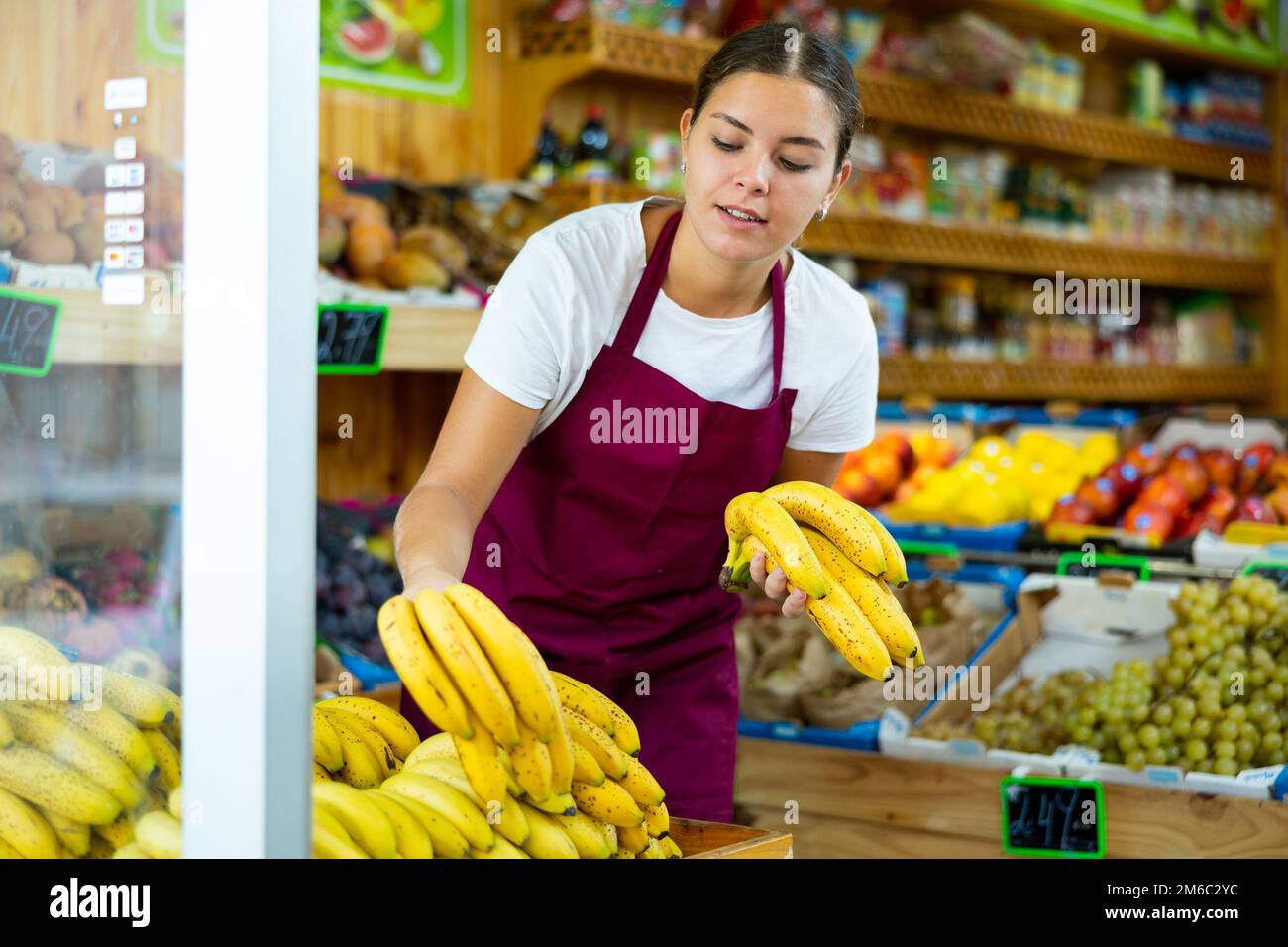 Smiling female merchandiser in uniform laying out bananas at grocery ...