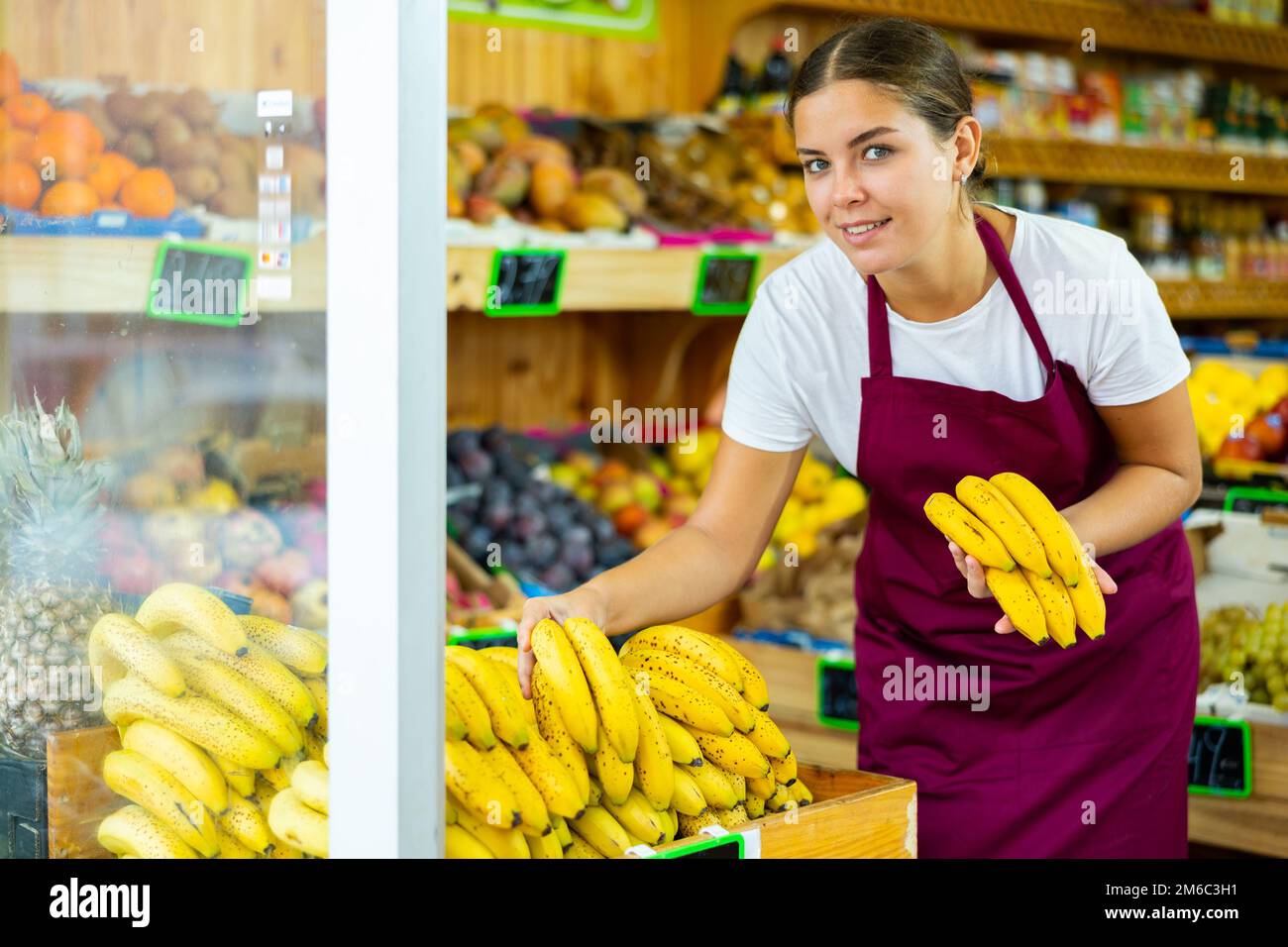 Grocery store female employee in an apron puts fresh bananas on shelves ...