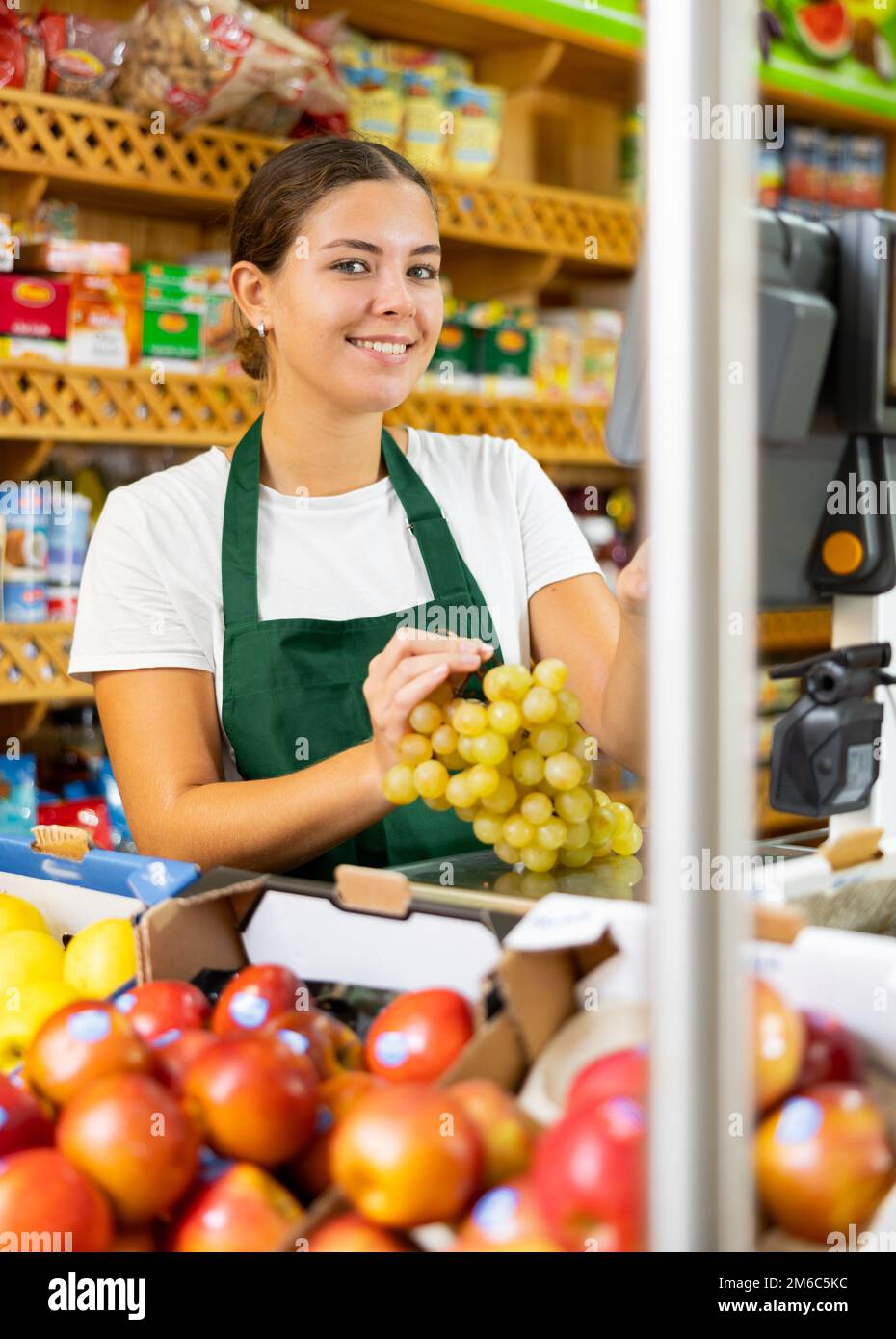 Friendly young female seller in apron weighing grape on scale during ...