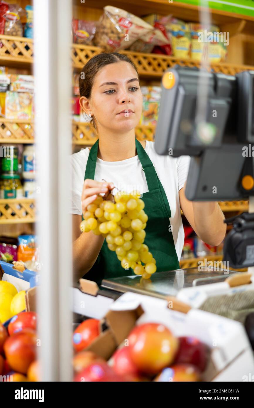 Young female supermarket worker weighing on the scales green grapes in ...