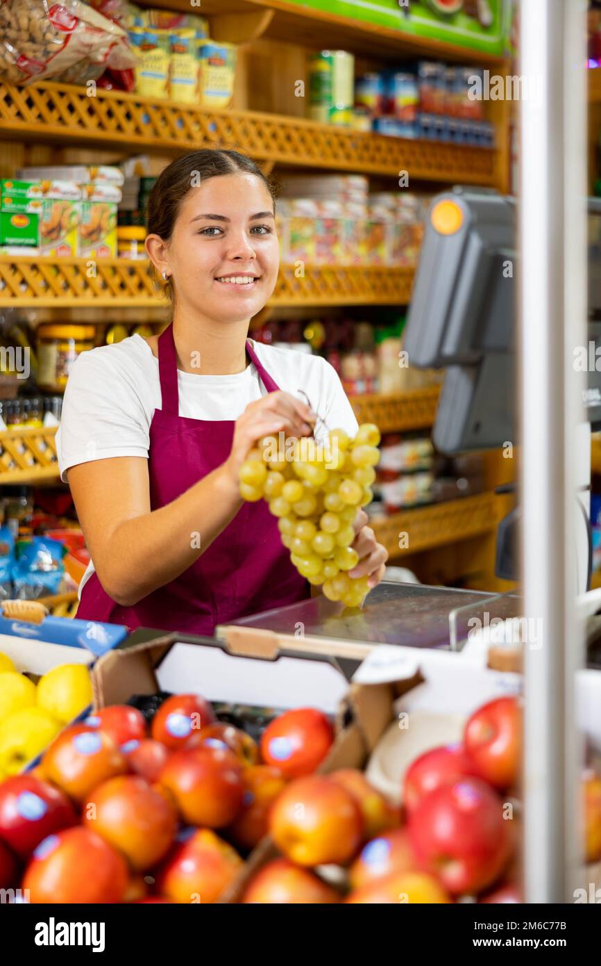 Positive young female supermarket worker in apron weighing on the ...