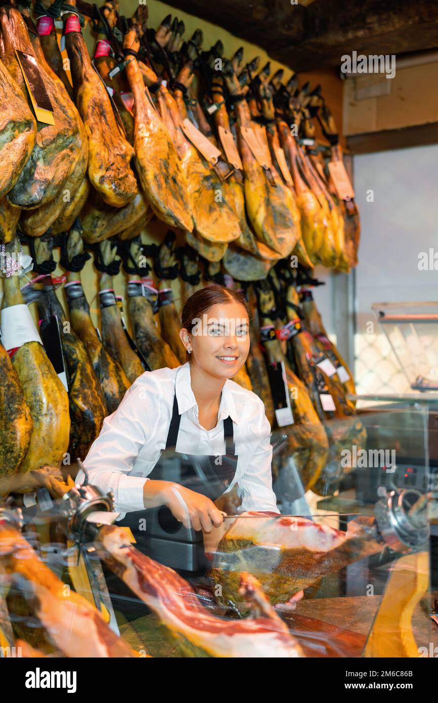 Female seller cutting slices from whole leg of jamon in meat store ...