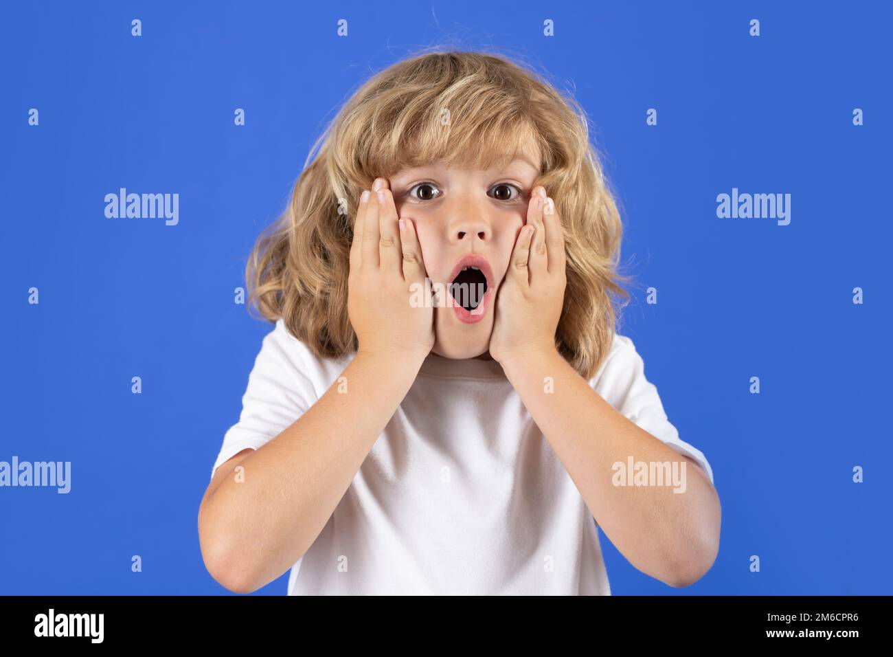 Shocked kid boy keeping hands near cheek with open mouth on blue ...