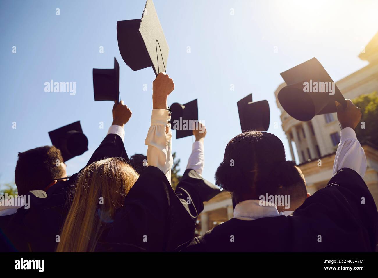 University graduates holding up their academic caps to bright blue sky ...