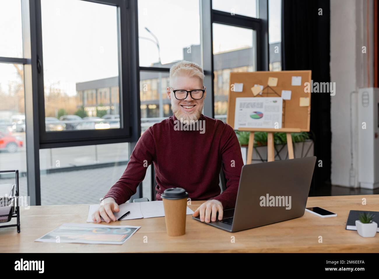 cheerful albino businessman in glasses looking at laptop near paper cup ...