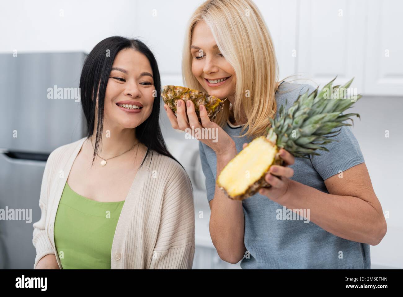 Smiling woman smelling fresh pineapple near asian friend in kitchen ...
