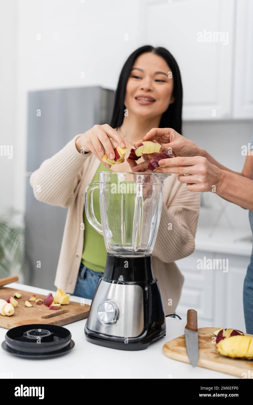 Woman putting apple in blender near blurred asian friend in kitchen ...