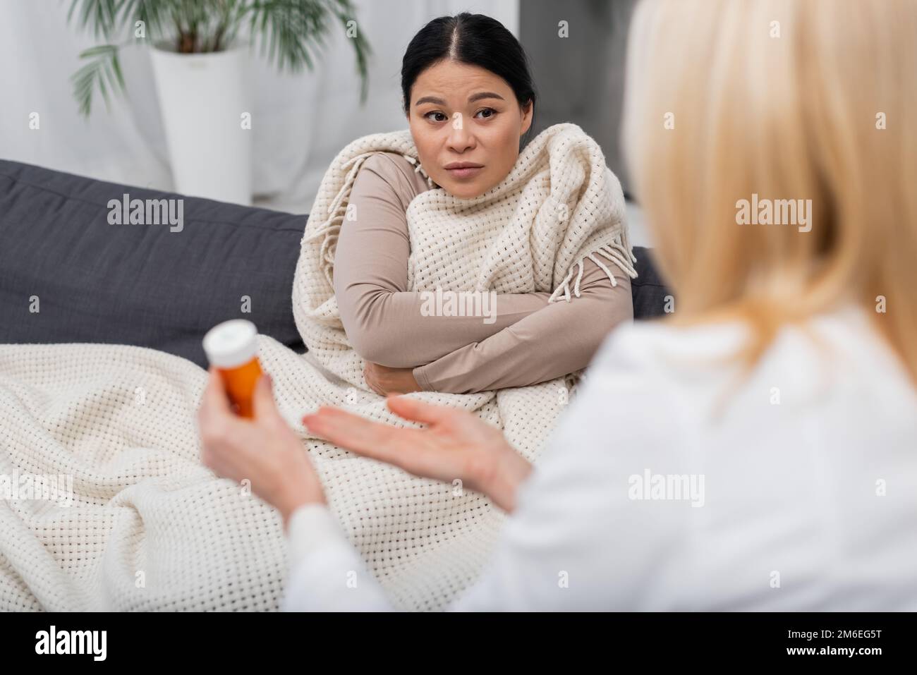 Sick asian woman in blanket looking at blurred doctor pointing at pills ...