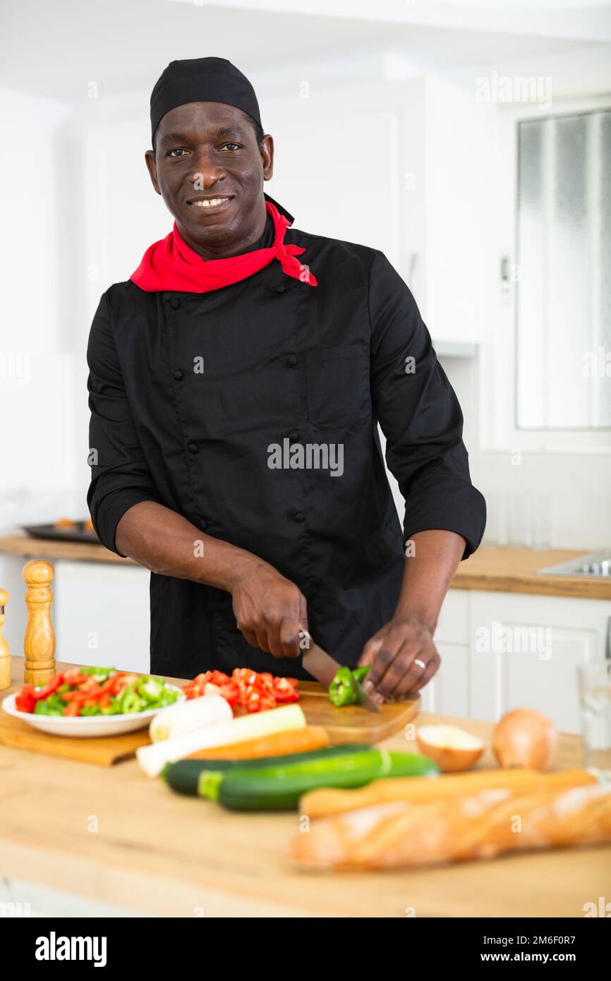 Cheerful chef in black uniform cutting vegetables for salad in home ...
