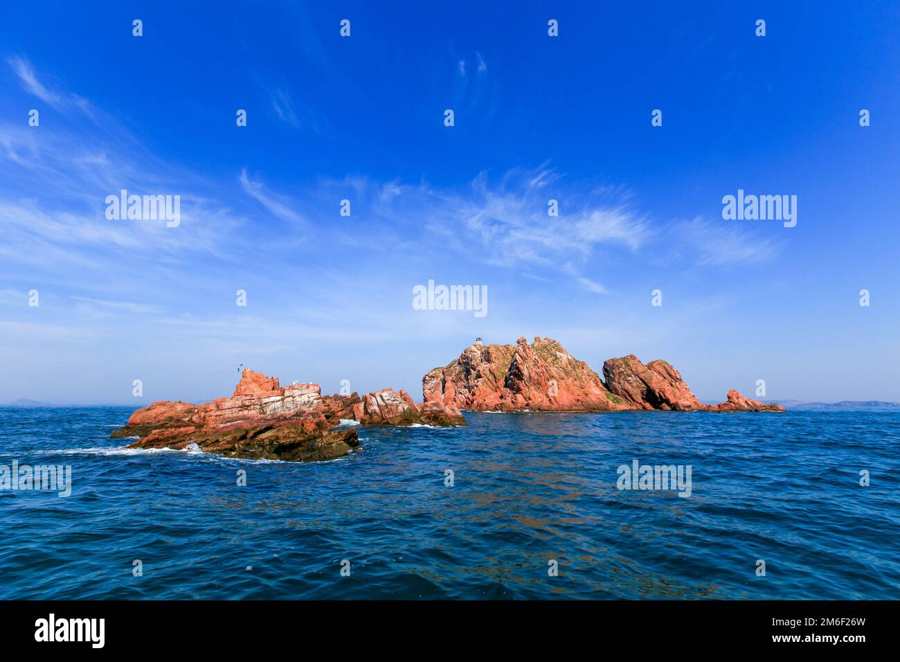 A picturesque red stone island sticks out of the blue sea water. An ...