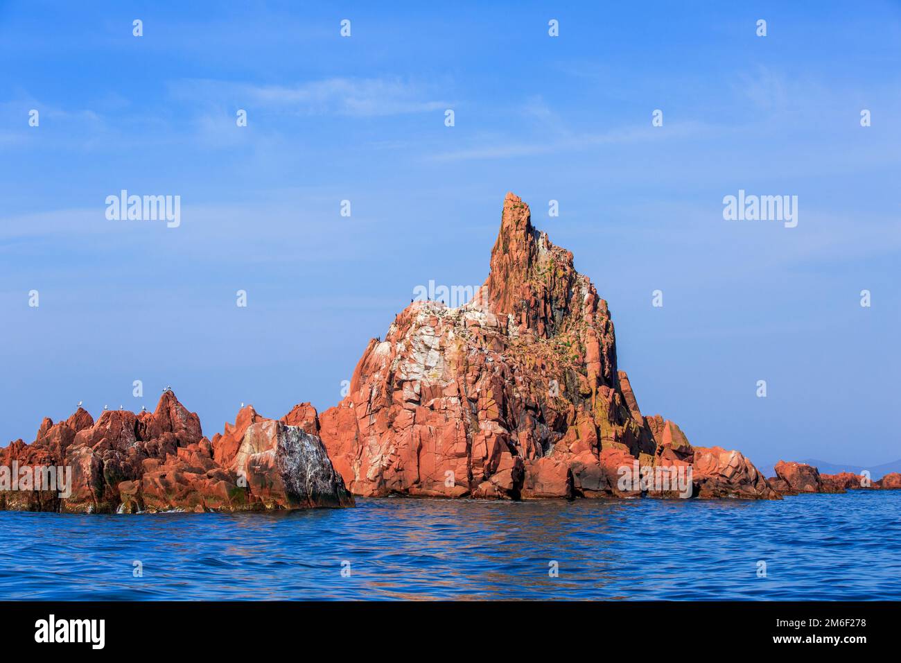 A picturesque red stone island sticks out of the blue sea water. An ...
