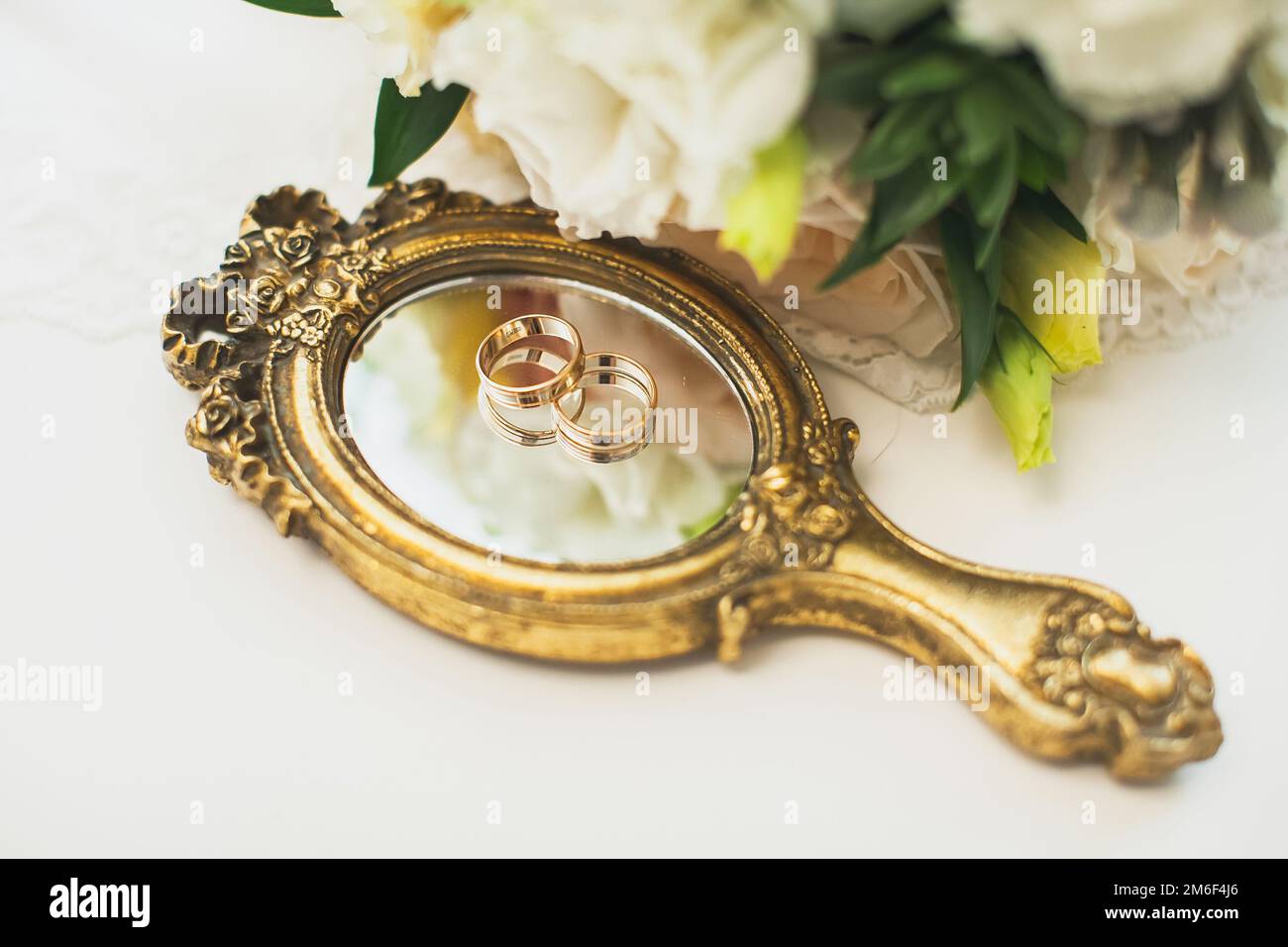 Wedding rings lie on mirror with a gilded handle. Nearby is a bouquet ...