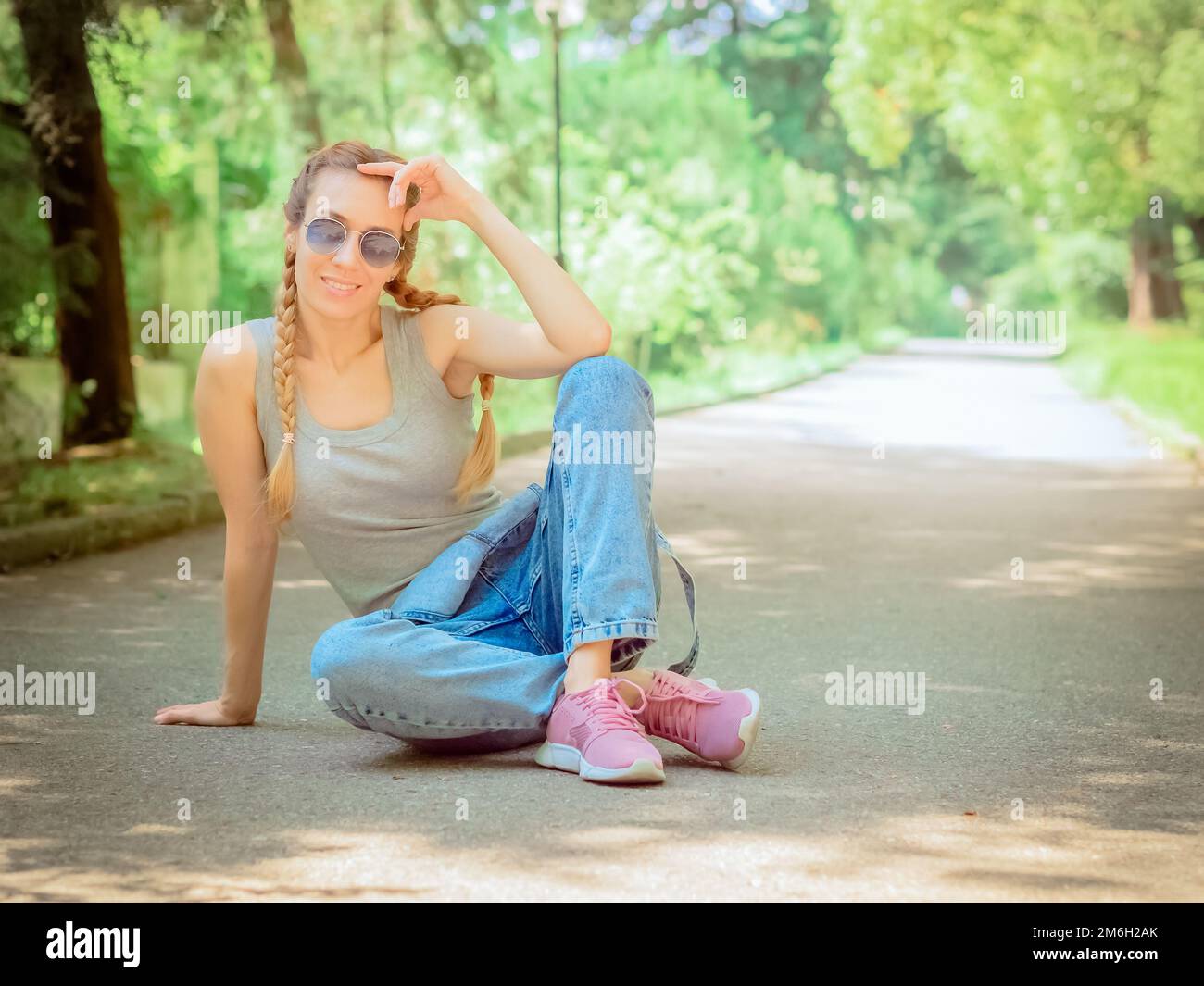 Smiling fair-haired girl with two pigtails sits on a paved path in a ...