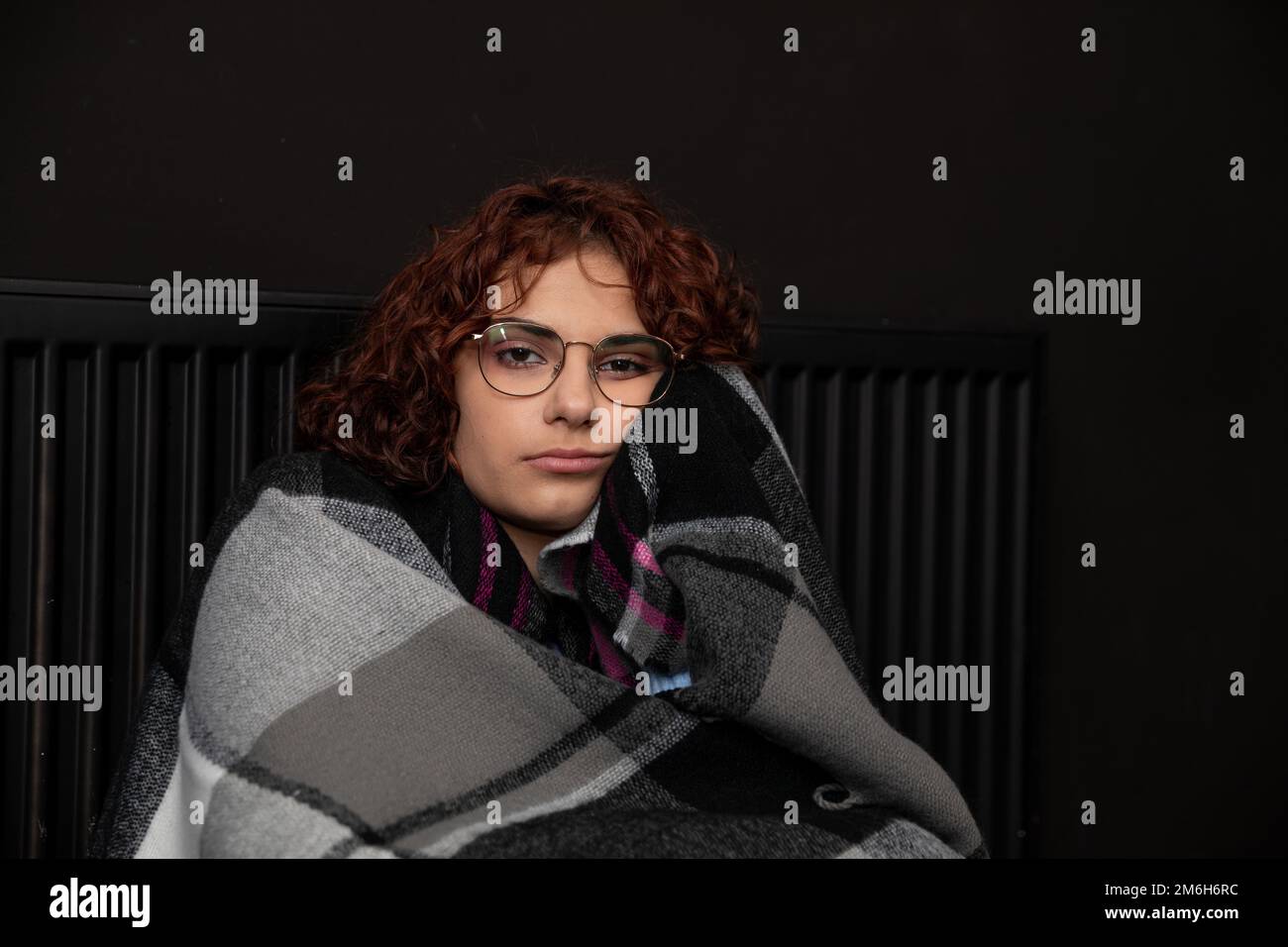 A sad and frozen girl covered with a thick scarf sits by the heater ...