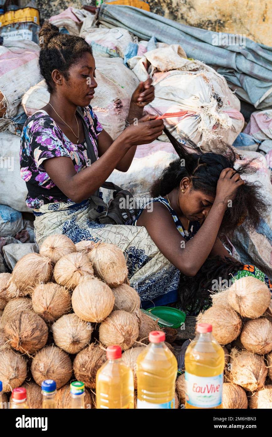 Two woman braiding hair, market stall, coconuts, bags of charcoal behind, Mahajanga, Madagascar ...