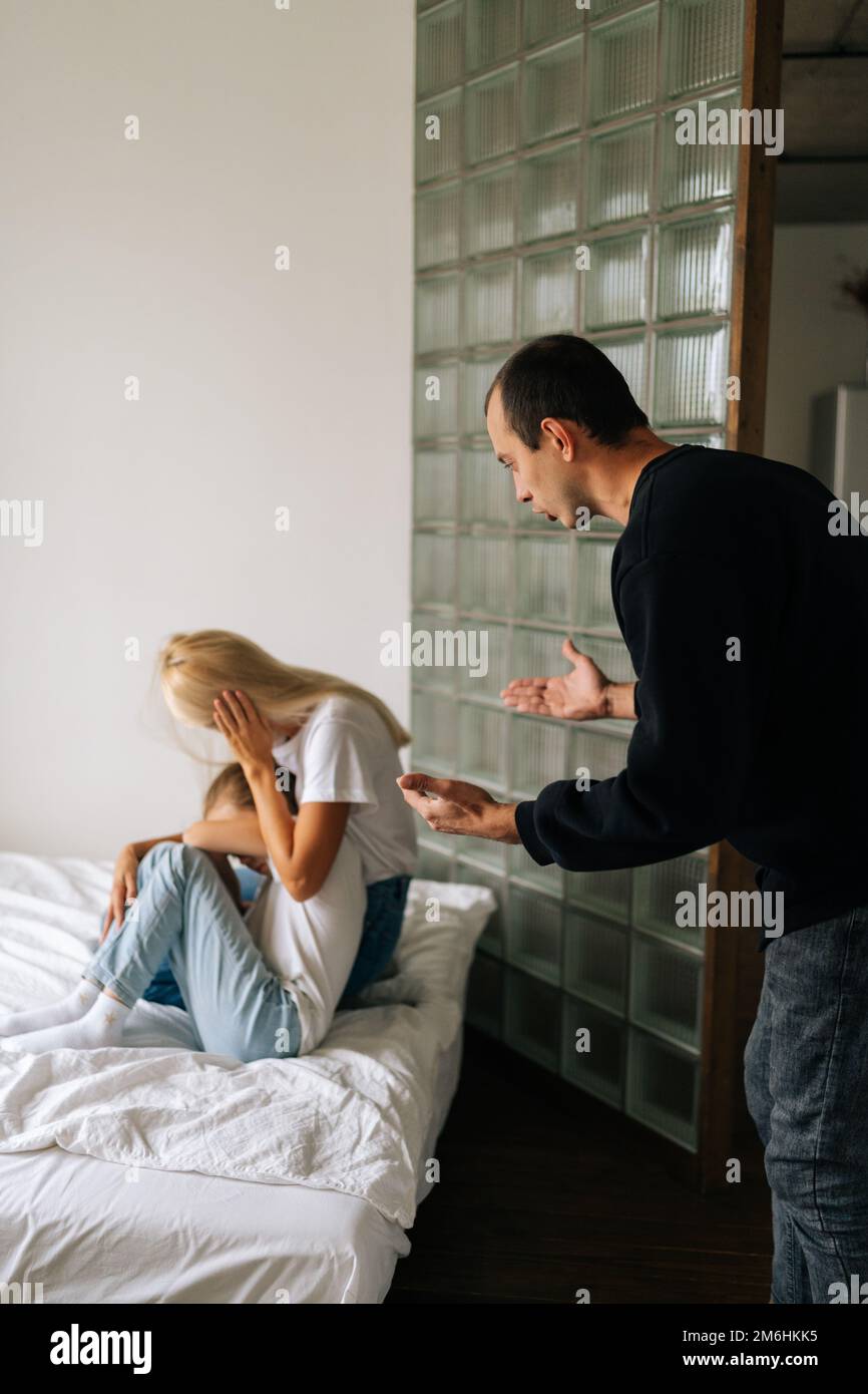 Portrait of scared mother and daughter hugging each other sitting on sofa while aggressive ...