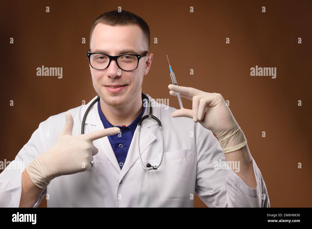 A young Caucasian doctor in a white coat and glasses holds a syringe ...