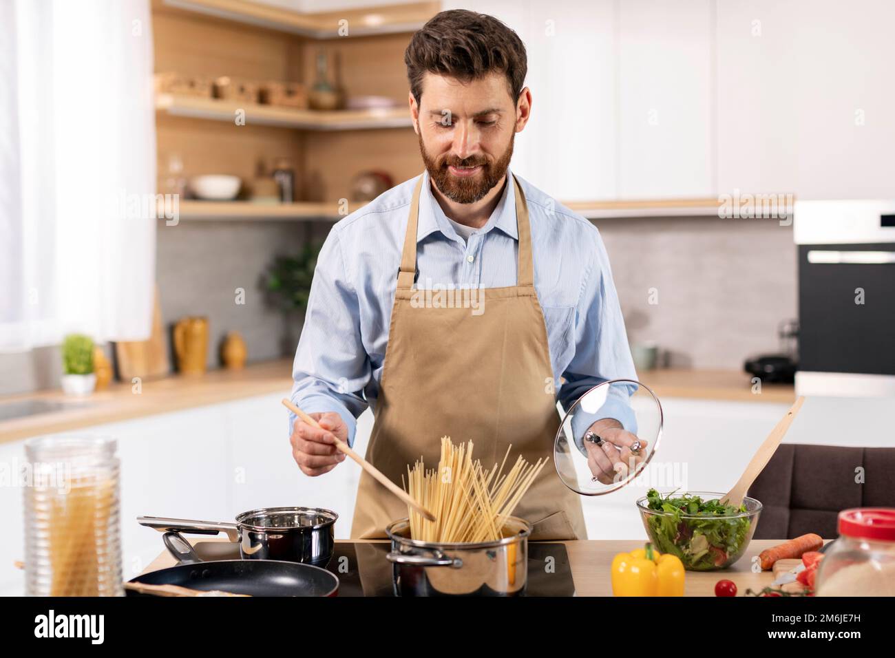 Smiling attractive millennial caucasian bearded guy in apron cooks ...