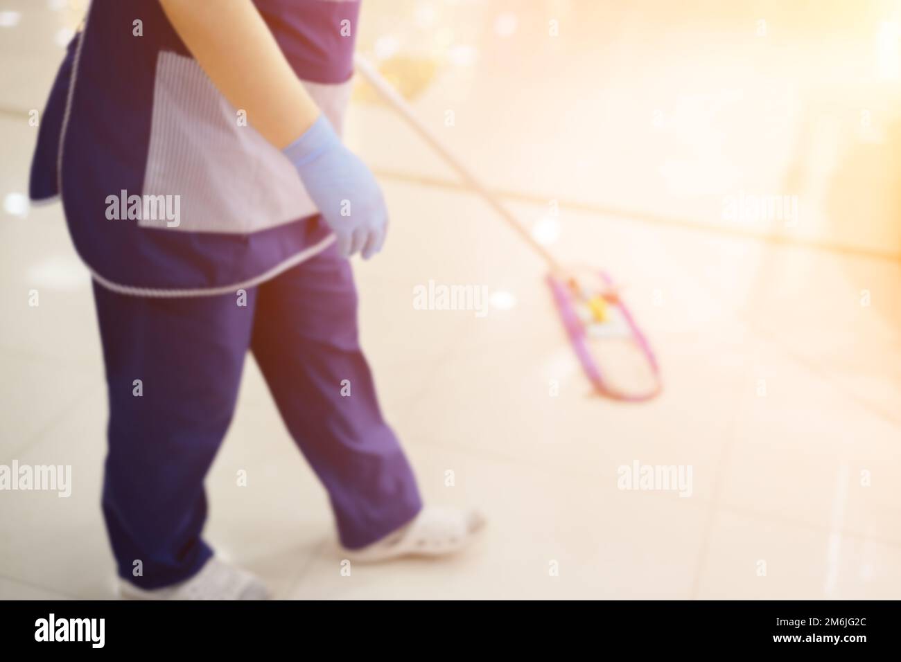 A cleaning lady in a blue uniform and gloves washes a tiled floor with ...