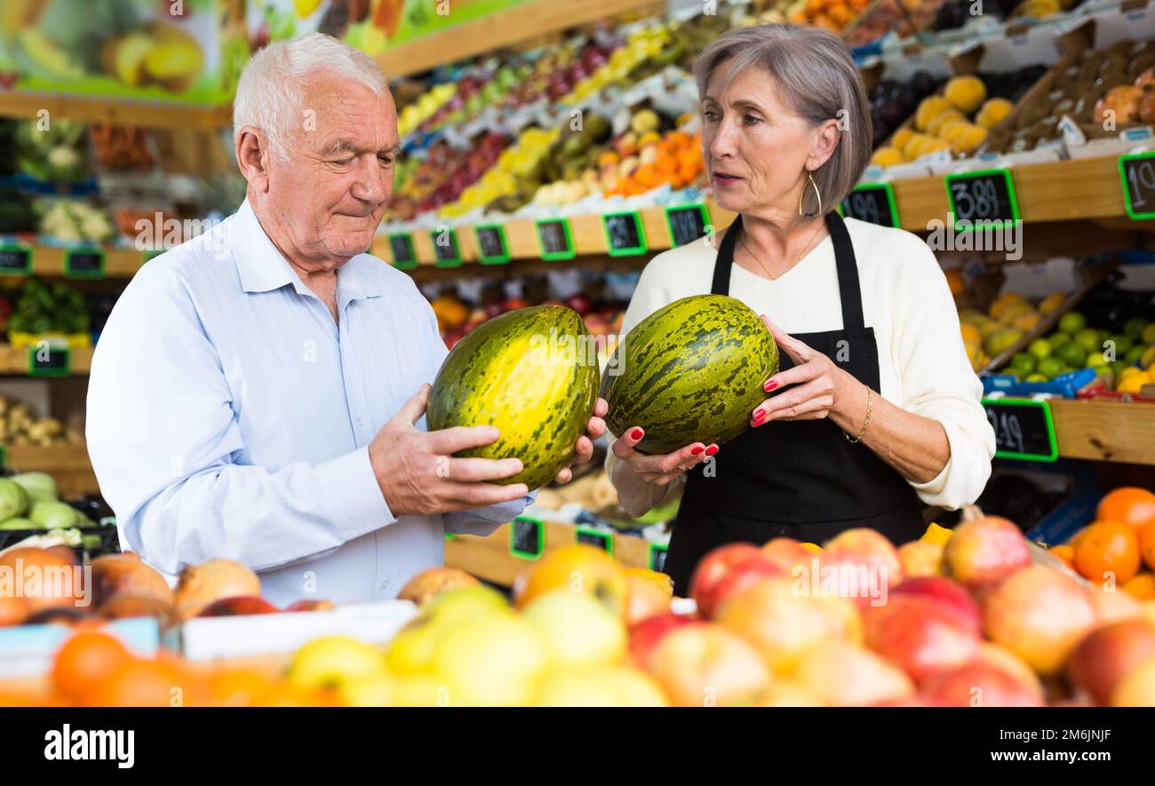 Woman supermarket employee helping mature man to choose watermelon and ...