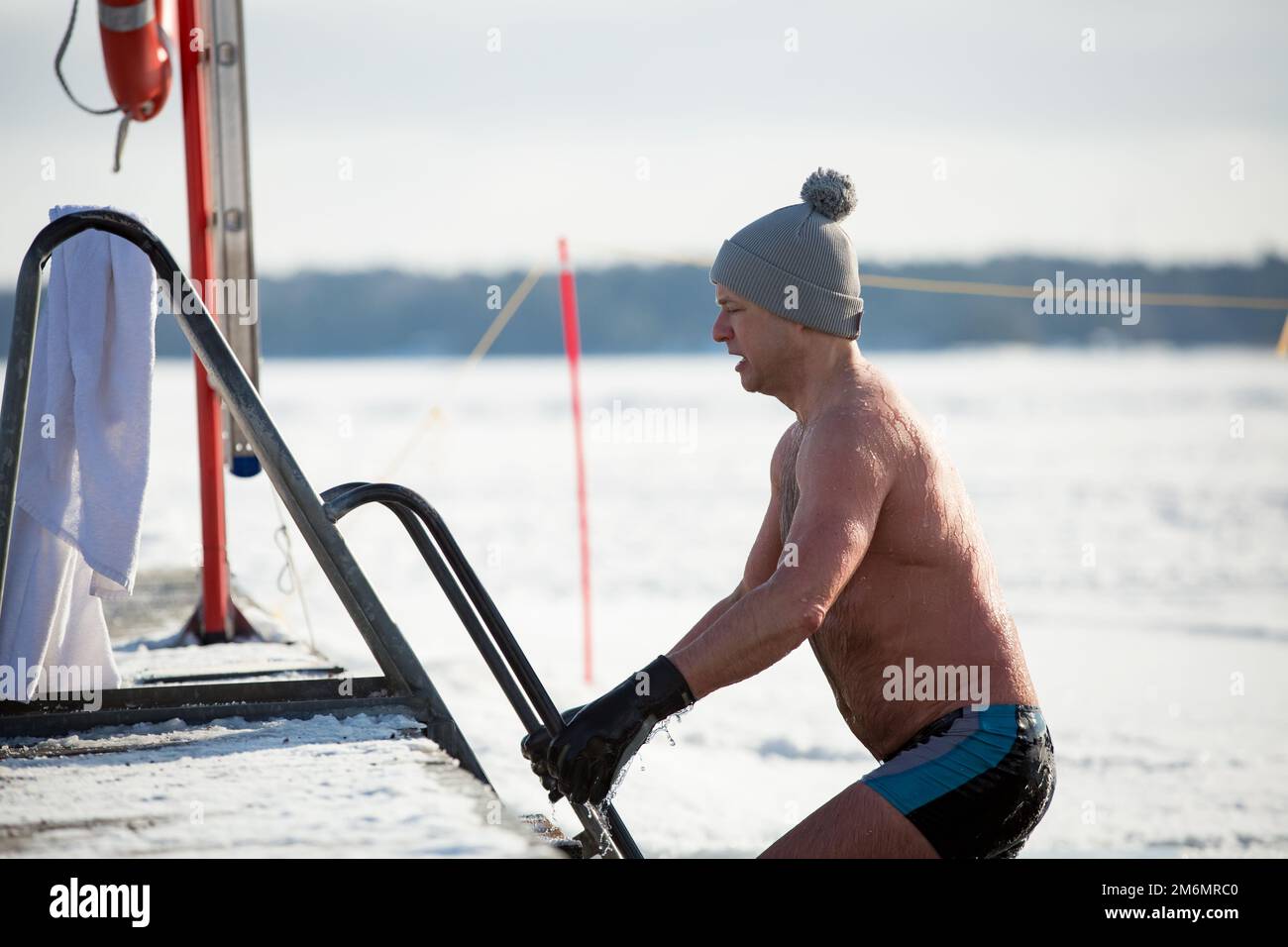 A man in warm hat and gloves swimming in an ice hole, coming out of the ...
