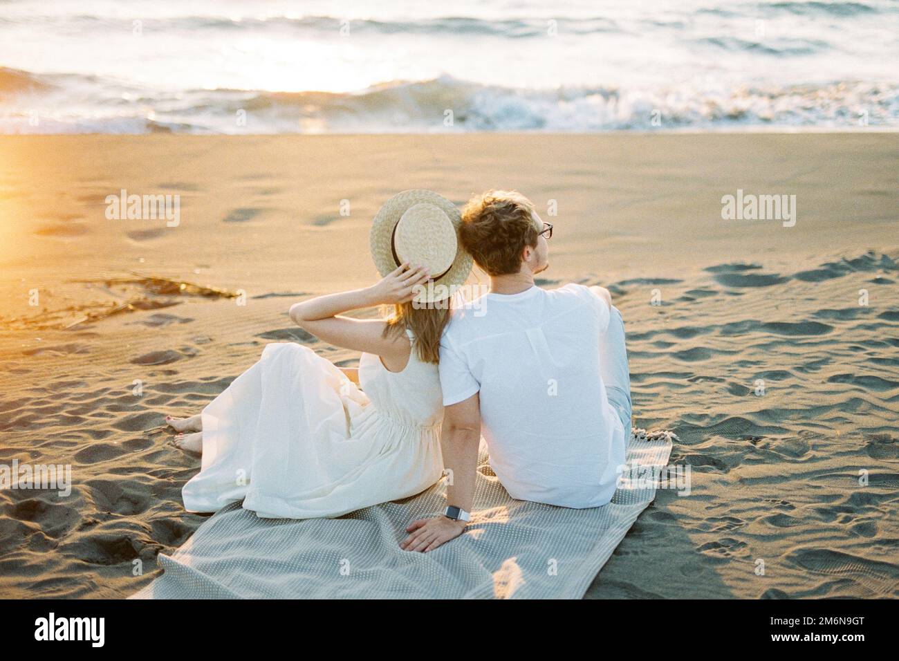 Woman in a hat sits on a blanket on the beach, leaning against a man ...