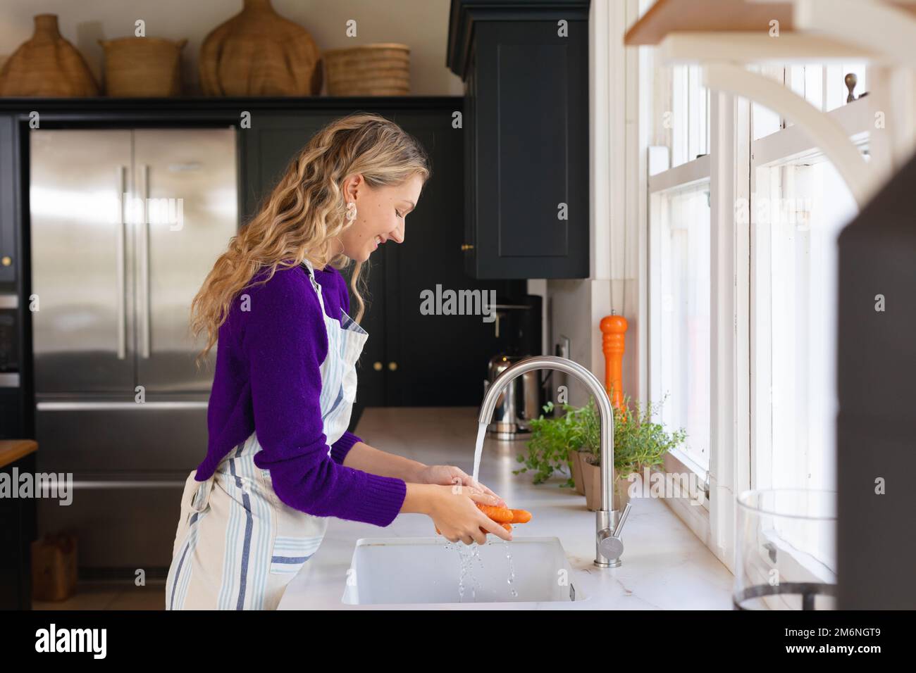 Smiling caucasian woman standing in kitchen in apron washing carrots in ...