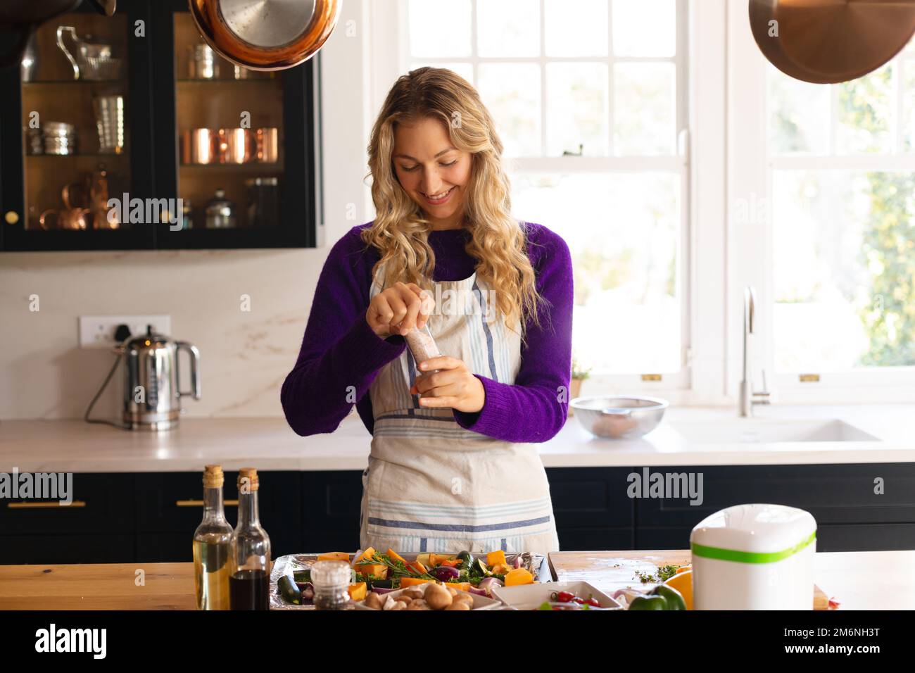 Smiling caucasian woman standing in kitchen in apron seasoning chopped ...