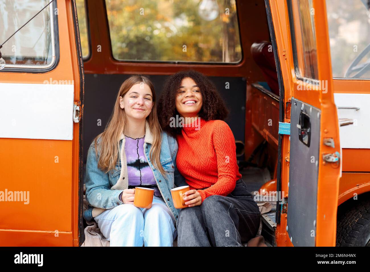 Two happy multiracial girlfriends sitting outdoors, drinking hot tea ...