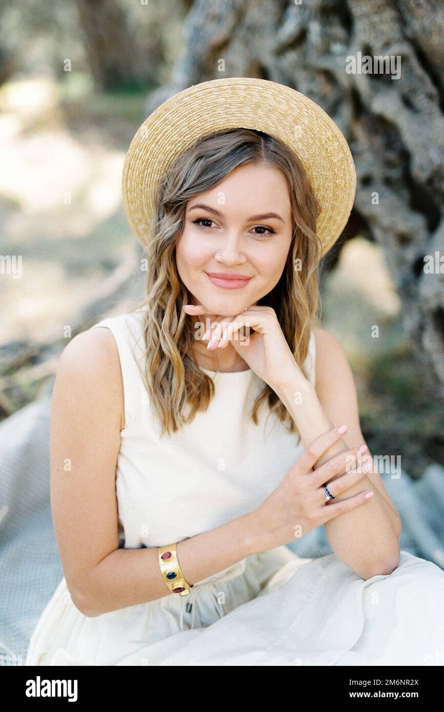 Young woman in a straw hat sits near a tree resting her chin on her hand Stock Photo - Alamy