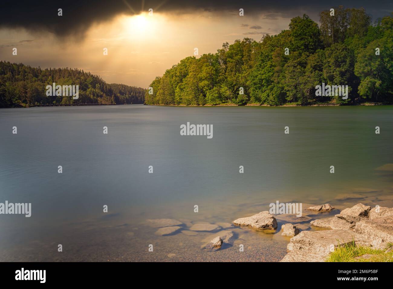 A closeup of a lake at sunset with trees around and sun on a cloudy sky ...