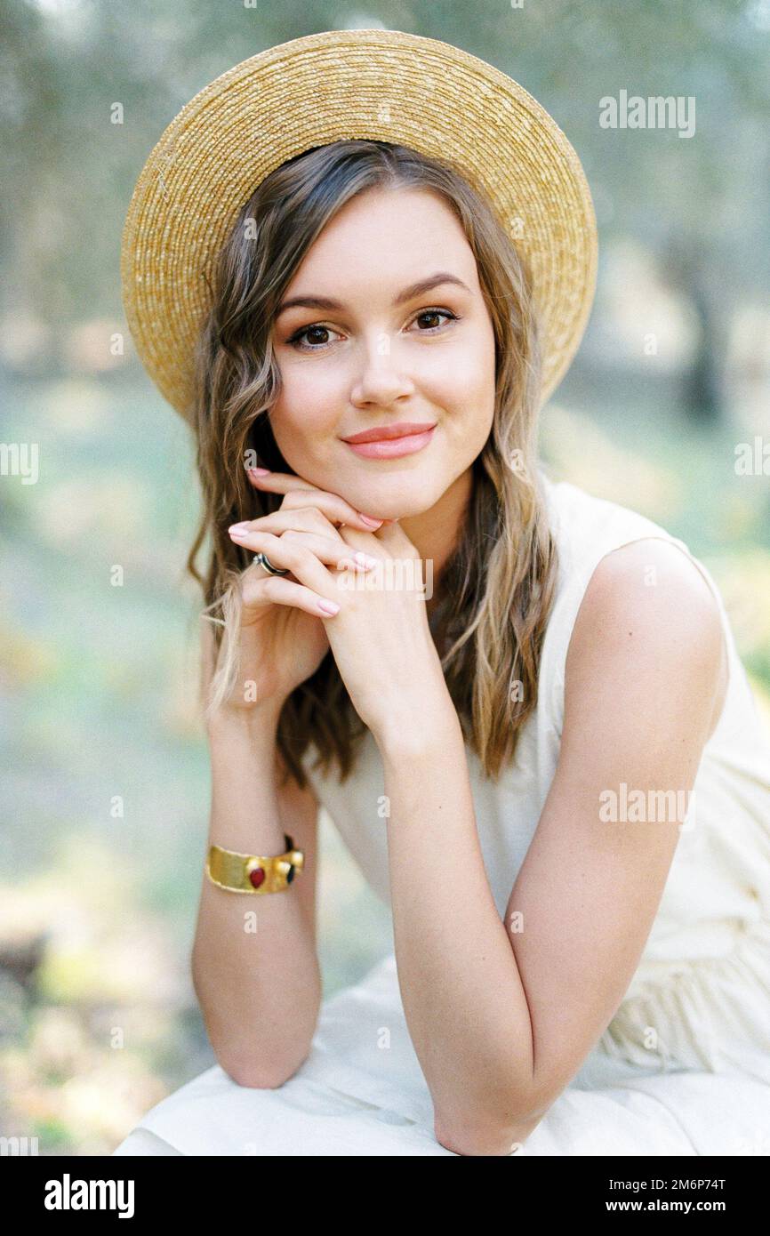 Young woman in a straw hat sits with her hands clasped under her chin ...