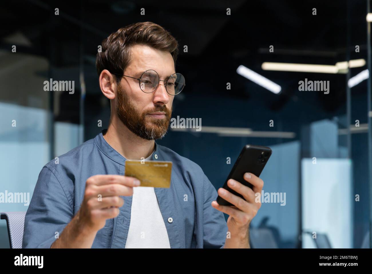 Serious thinking man inside office holding bank credit card and smartphone, businessman making ...