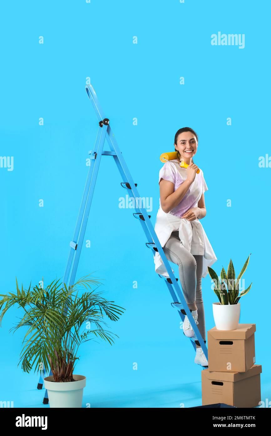 Young woman with paint roller, ladder and moving boxes on blue ...